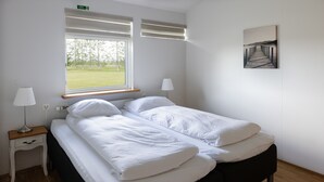 Uma cama de casal com lençóis brancos, uma mesa de cabeceira com abajur, uma janela com persianas e um quadro de um cais na parede.