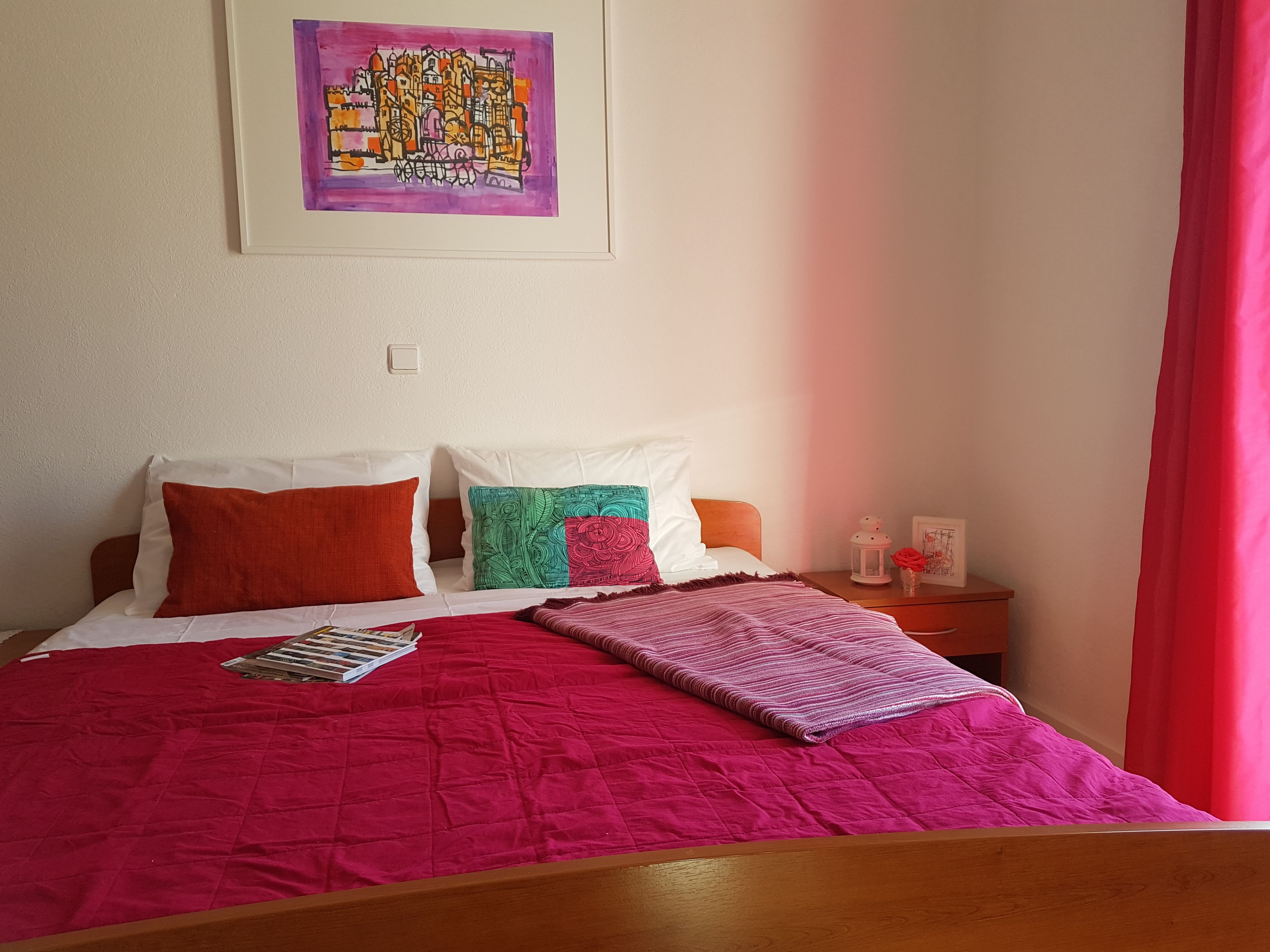 Cama bem arrumada com colcha vermelha, duas almofadas e um cobertor rosa. Uma moldura com um quadro está pendurada na parede acima da cama.