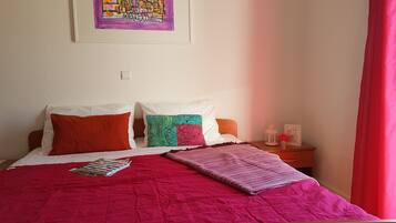 Cama bem arrumada com colcha vermelha, duas almofadas e um cobertor rosa. Uma moldura com um quadro está pendurada na parede acima da cama.