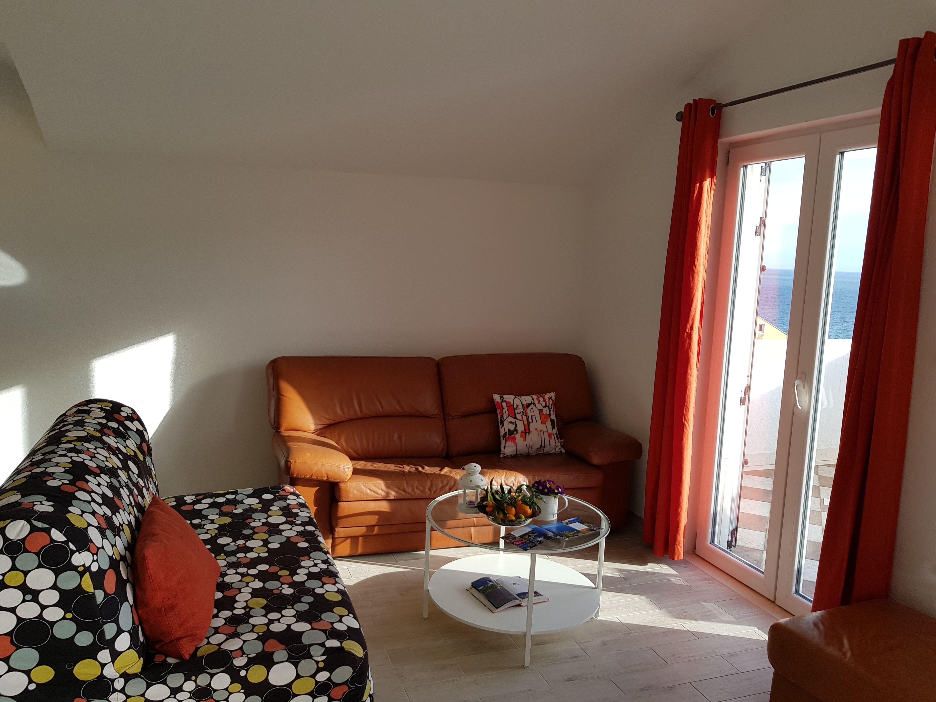 Sala de estar com sofá, mesa de centro com revistas e uma planta, além de vista através de uma porta de vidro.
