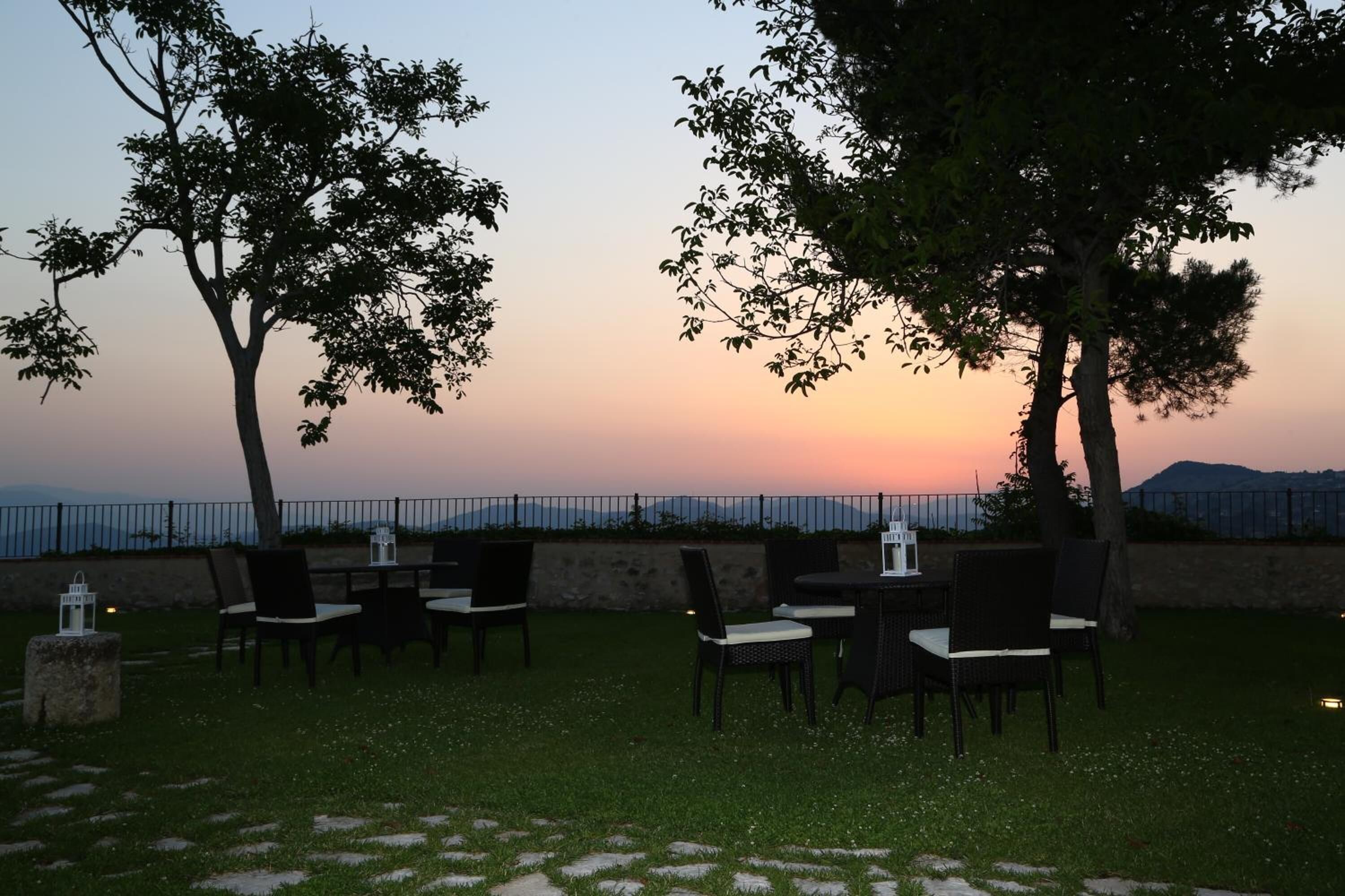 Uma área externa com mesas e cadeiras, uma parede de pedra e vista para um pôr do sol sobre a água.