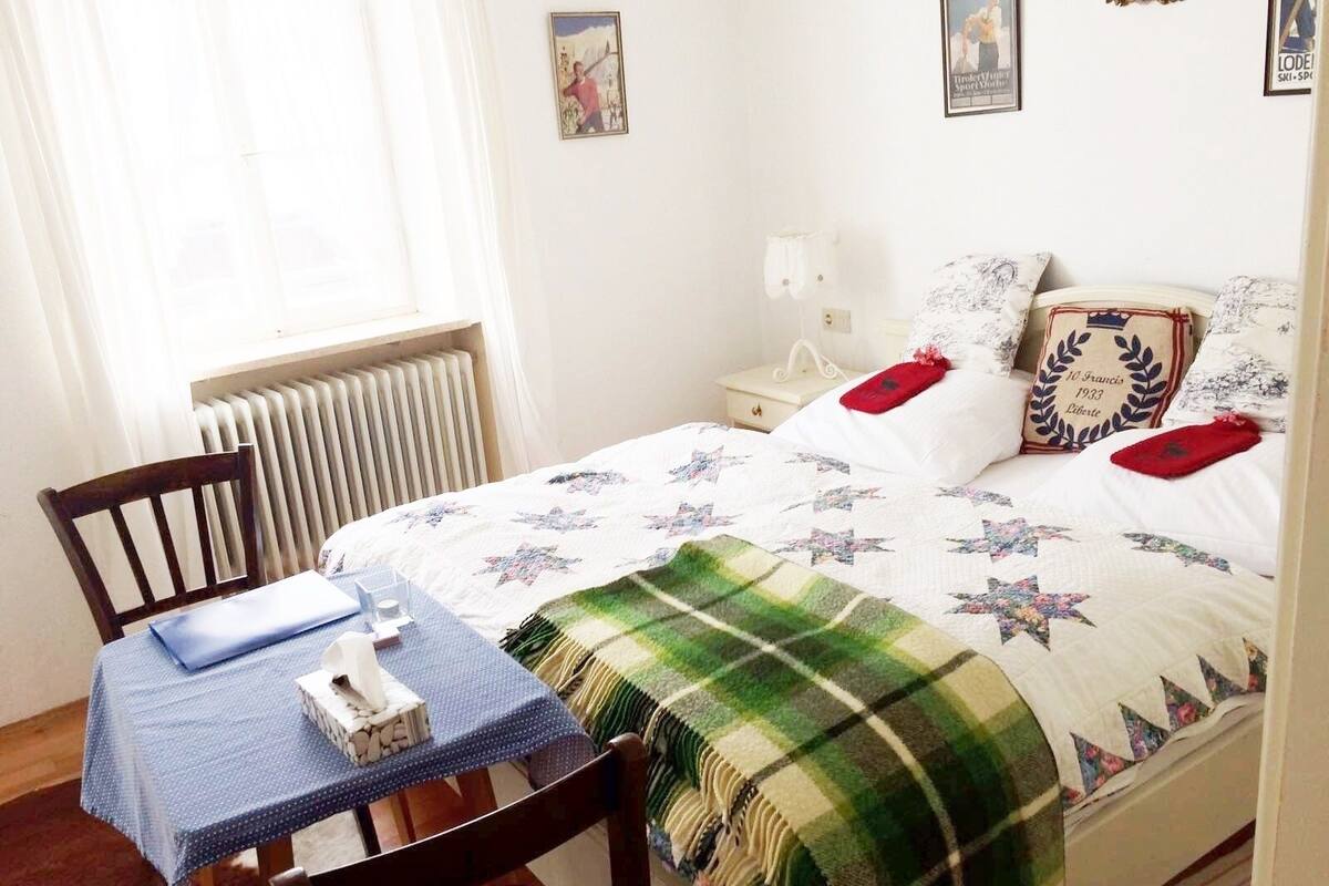 Quarto bem arrumado com cama, mesa de cabeceira, cadeira e radiador.