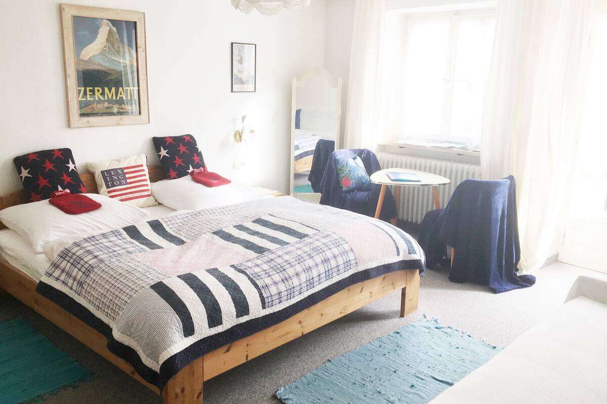 Quarto com cama de madeira, uma pequena mesa redonda e uma cadeira. Há uma janela com cortinas e um radiador.