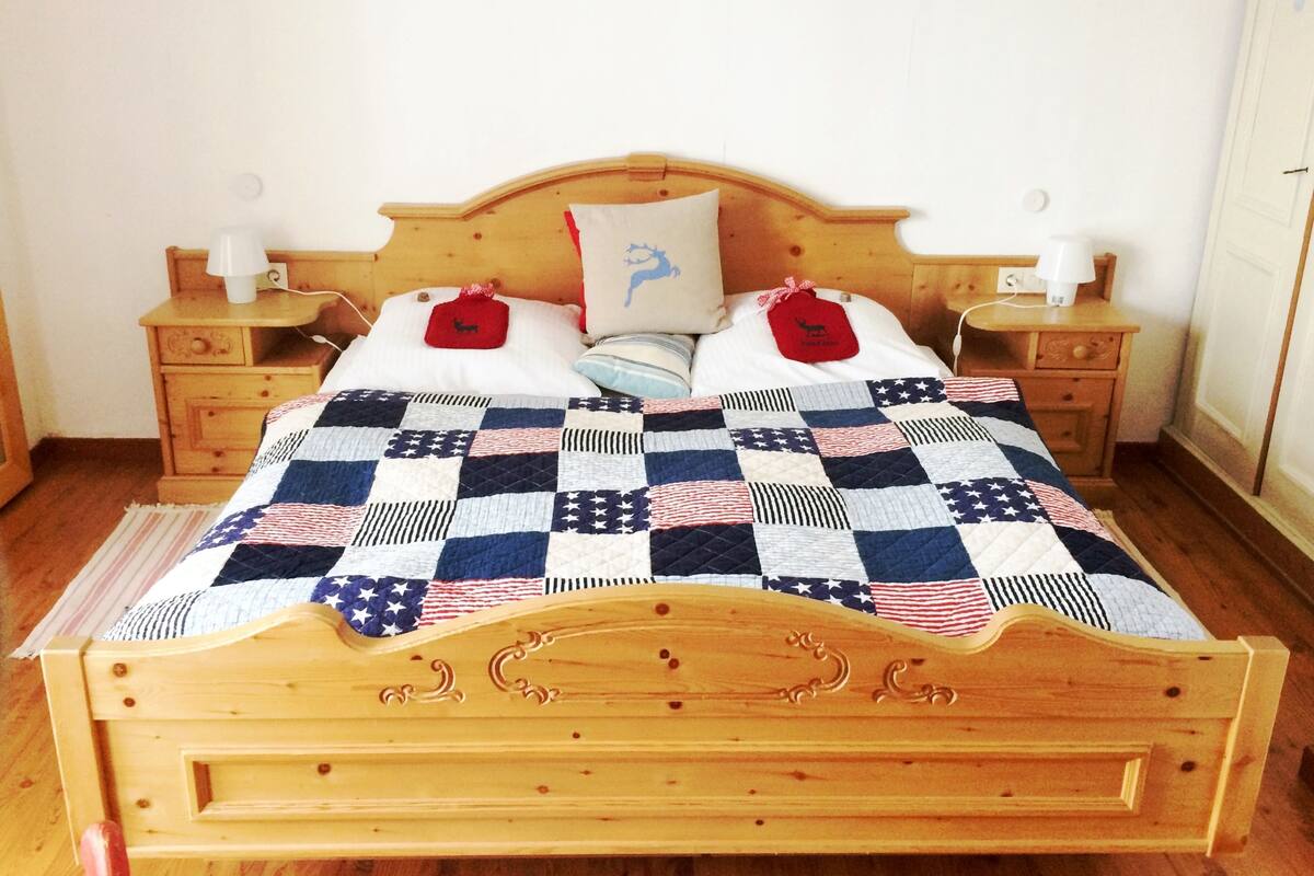 Uma cama de madeira com colcha estampada em xadrez, duas mesas de cabeceira com abajures e uma mesinha de apoio com uma luminária.