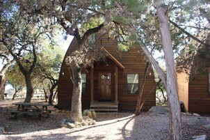 Uma cabana rústica com fachada de madeira, varanda frontal e mesa de piquenique numa área arborizada.