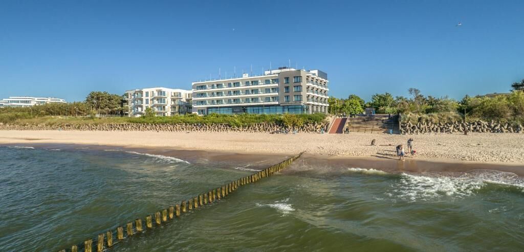 Um hotel à beira-mar com vários andares, uma praia arenosa ao fundo e uma barreira de madeira a separar a praia da água.