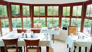 Uma sala de jantar com mesas preparadas para o café da manhã, janelas amplas com vista para o verde e um ambiente acolhedor.