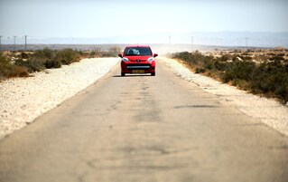 Um carro vermelho percorre uma estrada de cascalho com vegetação rasteira em ambos os lados.