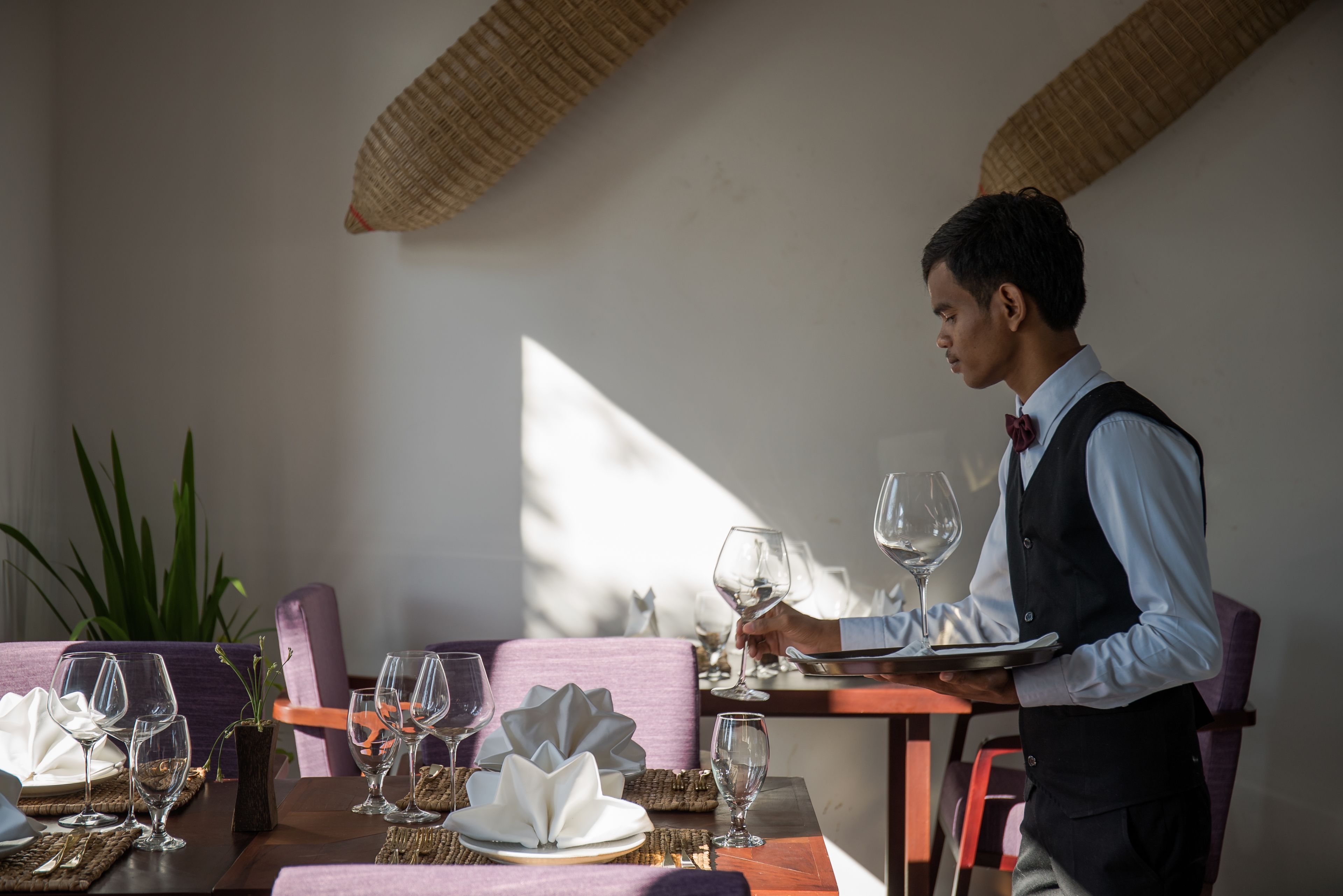 Garçom servindo vinho numa mesa com uma cadeira roxa e uma planta ao fundo.