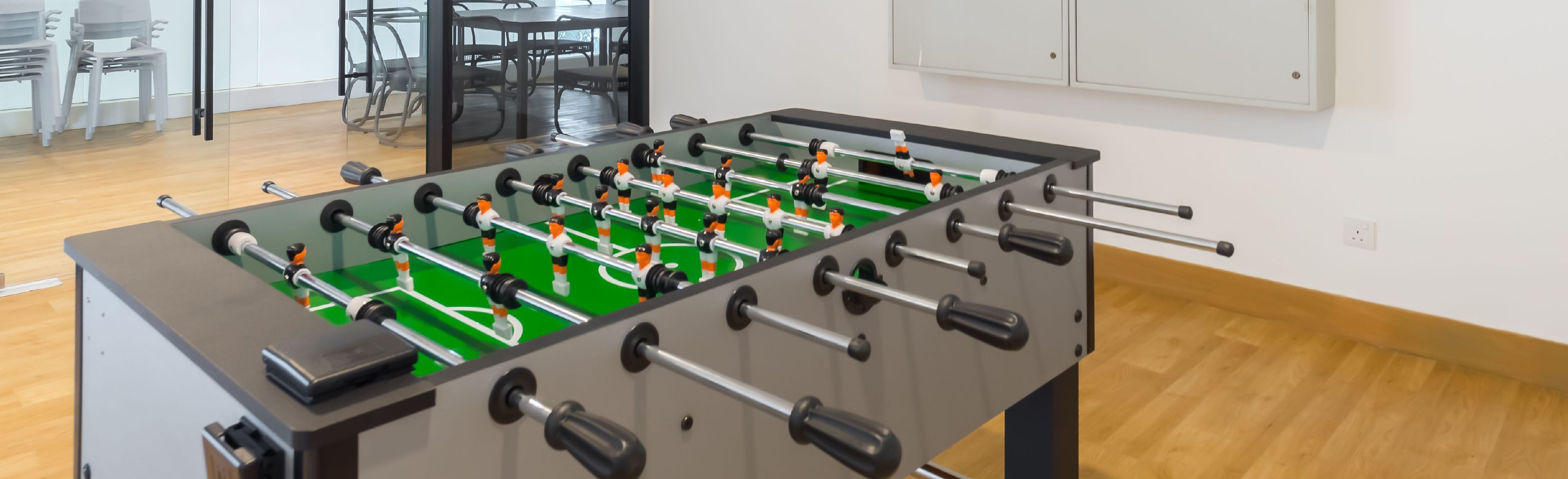 Uma mesa de futebol de mesa em um quarto com piso de madeira e cadeiras ao fundo.