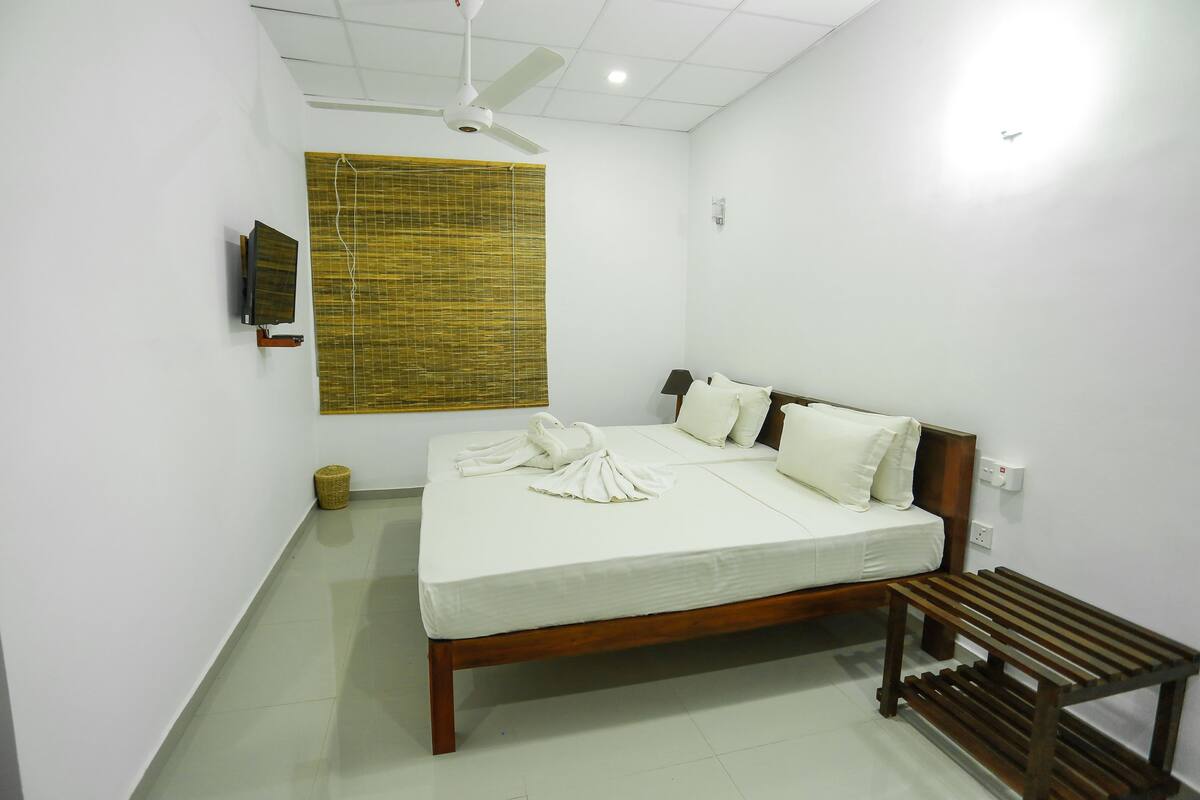 Quarto com cama, um banco de madeira, ventoinha no teto, televisão na parede e janela com persianas.