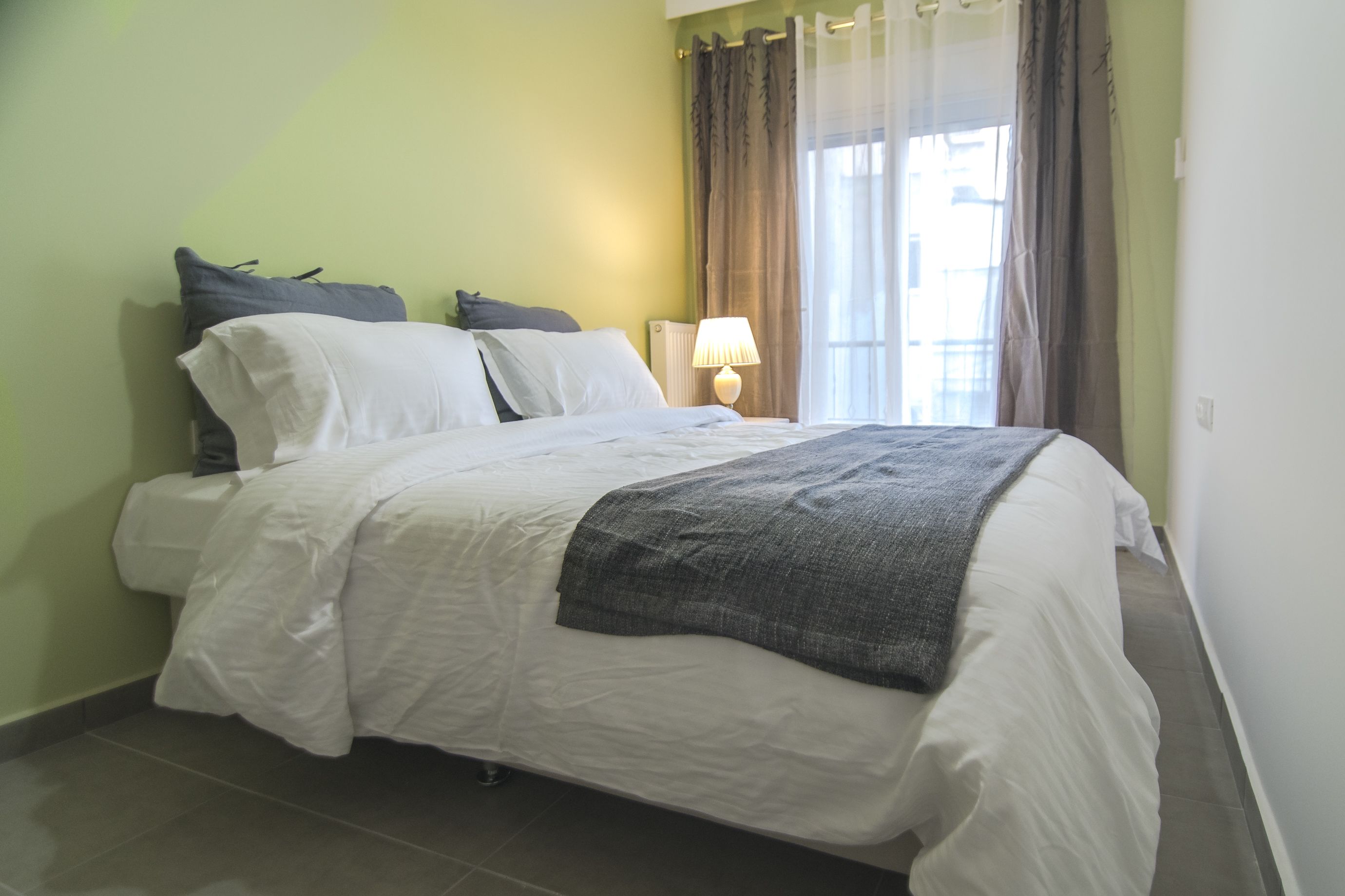 Cama bem arrumada com roupa de cama branca e manta cinza, uma luminária de cabeceira e uma janela com cortinas.