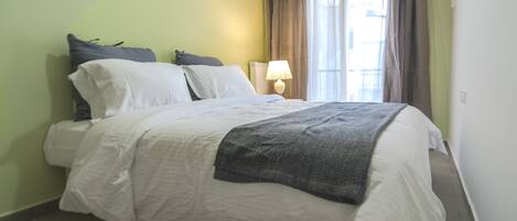 Cama bem arrumada com roupa de cama branca e manta cinza, uma luminária de cabeceira e uma janela com cortinas.