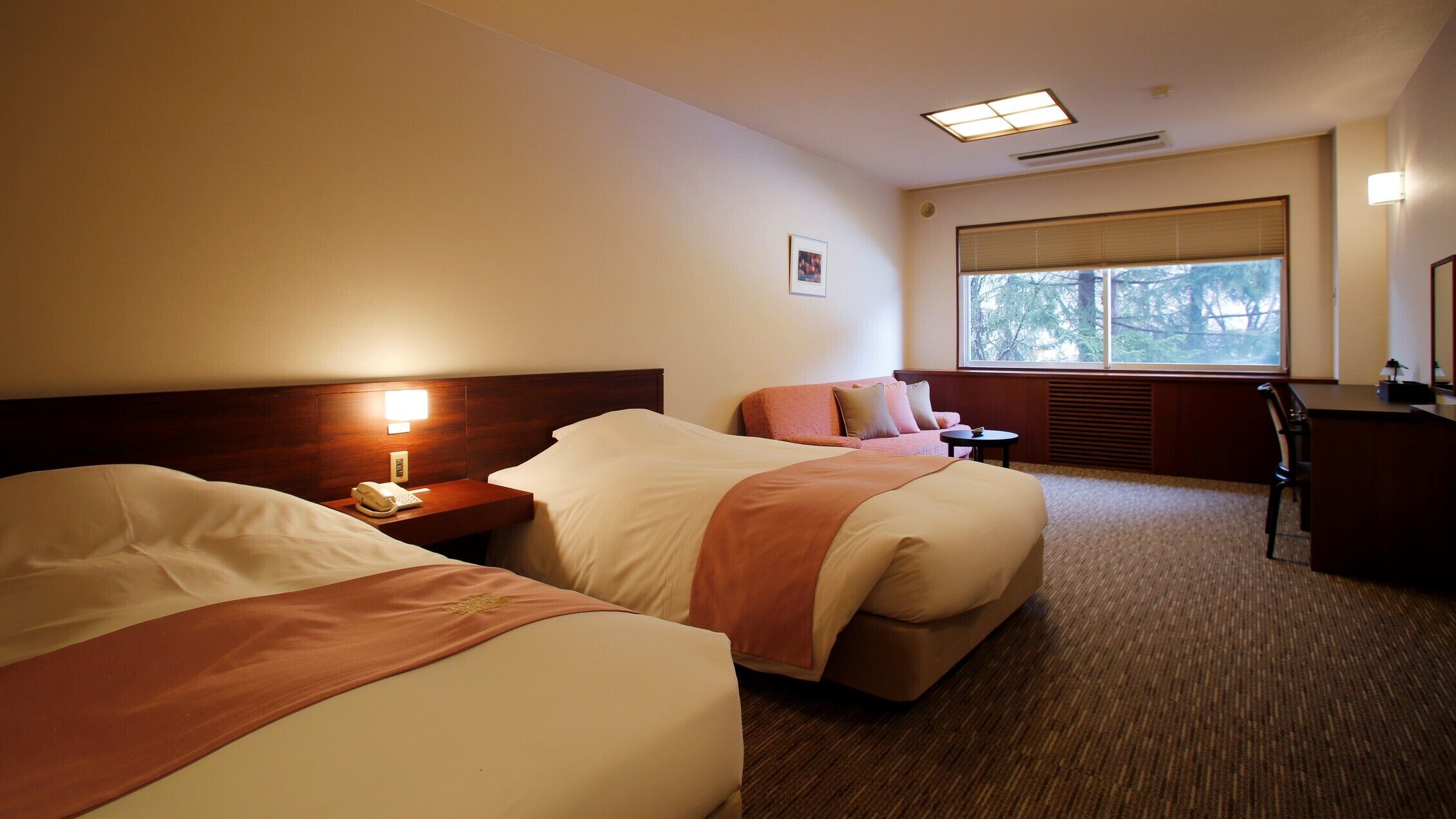 Quarto de hotel com duas camas, uma mesa, uma cadeira, uma janela com vista e um abajur.