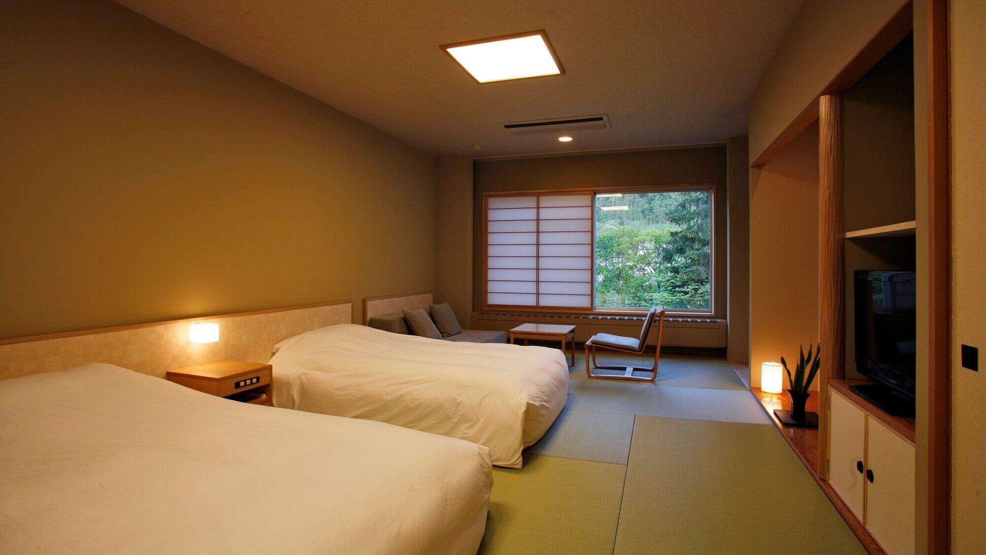 Quarto de hotel com duas camas, uma mesa pequena, uma cadeira, televisão e uma janela grande com vista para o verde.