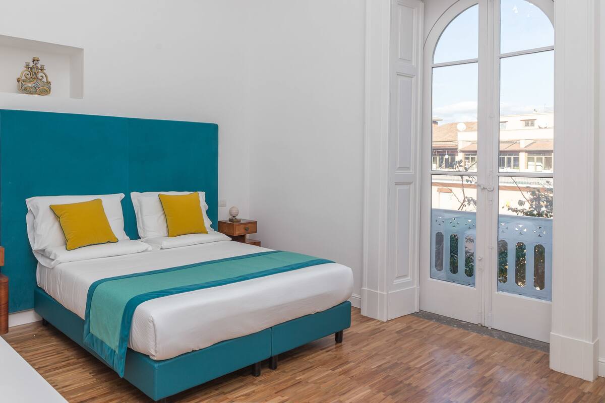 Quarto com cama grande, duas mesas de cabeceira, janela com vista e objetos decorativos na parede.