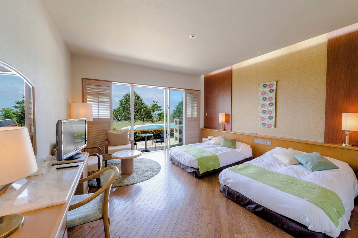 Quarto de hotel com duas camas, uma escrivaninha com computador, uma cadeira e vista para o exterior.