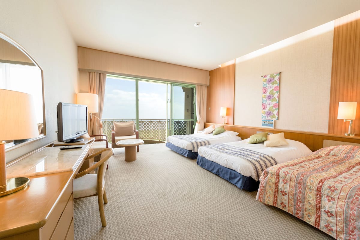 Quarto de hotel com duas camas, uma escrivaninha com televisão, uma cadeira e varanda com vista.