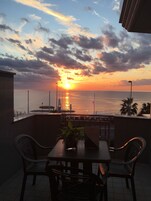 Terraço com mesa e cadeiras, com vista para o pôr do sol sobre o mar.