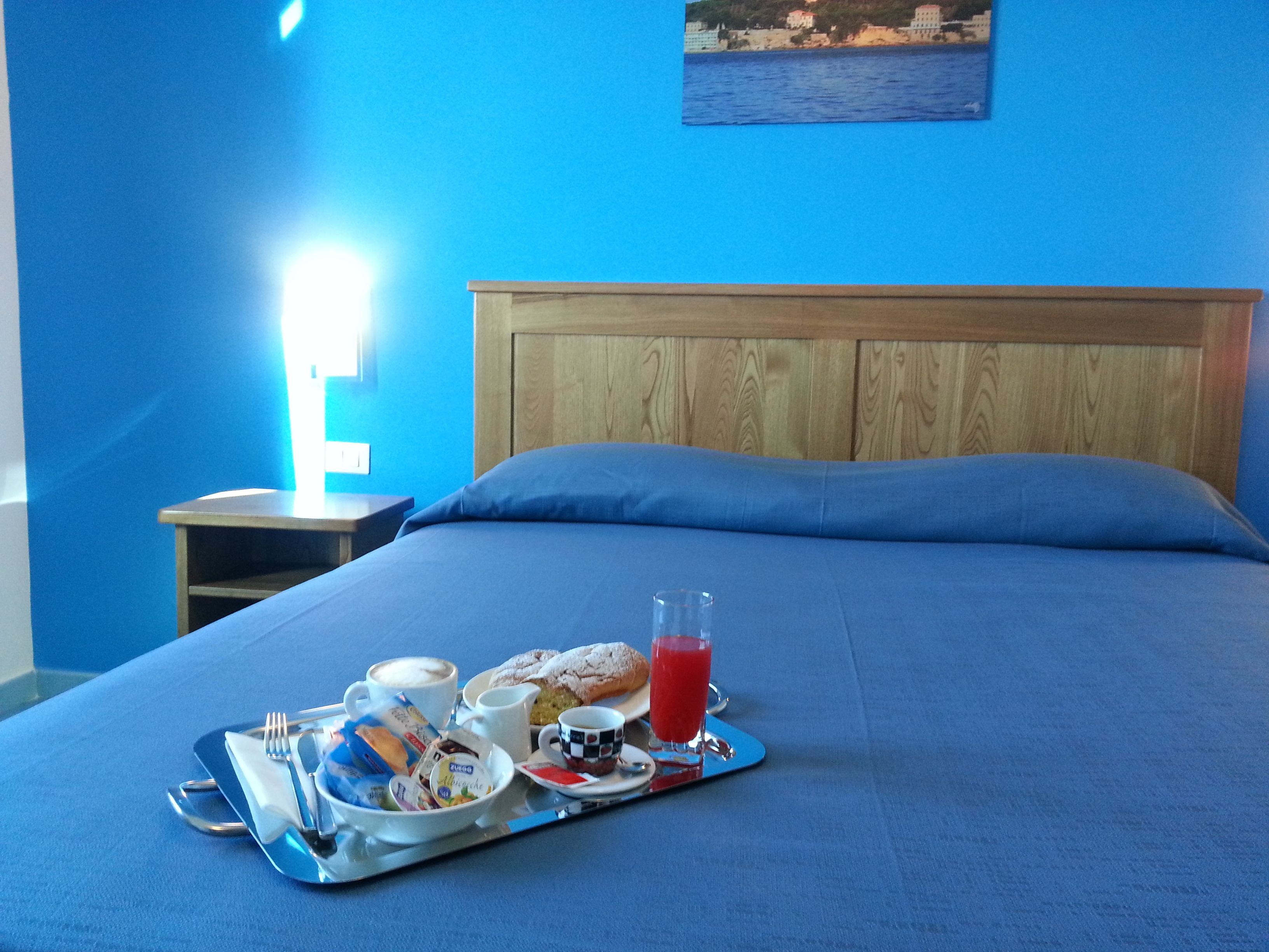 Cama bem arrumada com colcha azul, mesa de cabeceira com abajur e bandeja com itens de café da manhã, incluindo um pastel, café e suco.