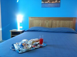 Cama bem arrumada com colcha azul, mesa de cabeceira com abajur e bandeja com itens de café da manhã, incluindo um pastel, café e suco.