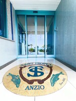 Entrada de hotel com porta de vidro, uma placa com a inscrição 'SAINT TROPEZ ANZIO' e um emblema circular com um cifrão e golfinhos.