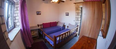 Quarto com cama, cabeceira de madeira, ventilador de teto, janela com cortinas e uma pequena mesa de madeira.