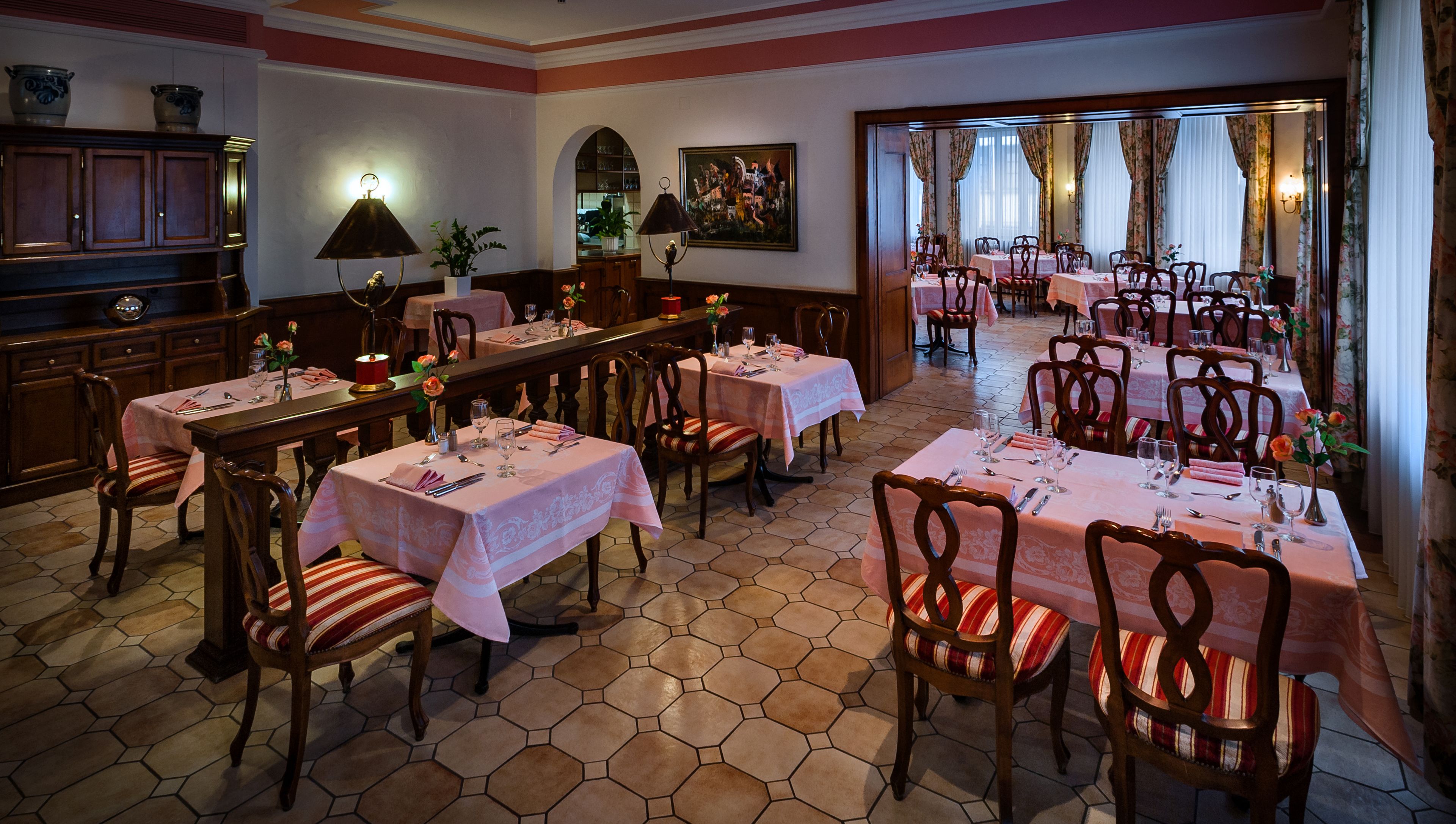 Um restaurante com chão de azulejos hexagonais, cadeiras de madeira com almofadas listradas e mesas dispostas com toalhas brancas e talheres.