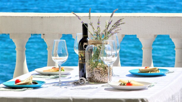 Uma mesa posta com pratos, copos, uma garrafa e um vaso de flores, com vista para o mar.