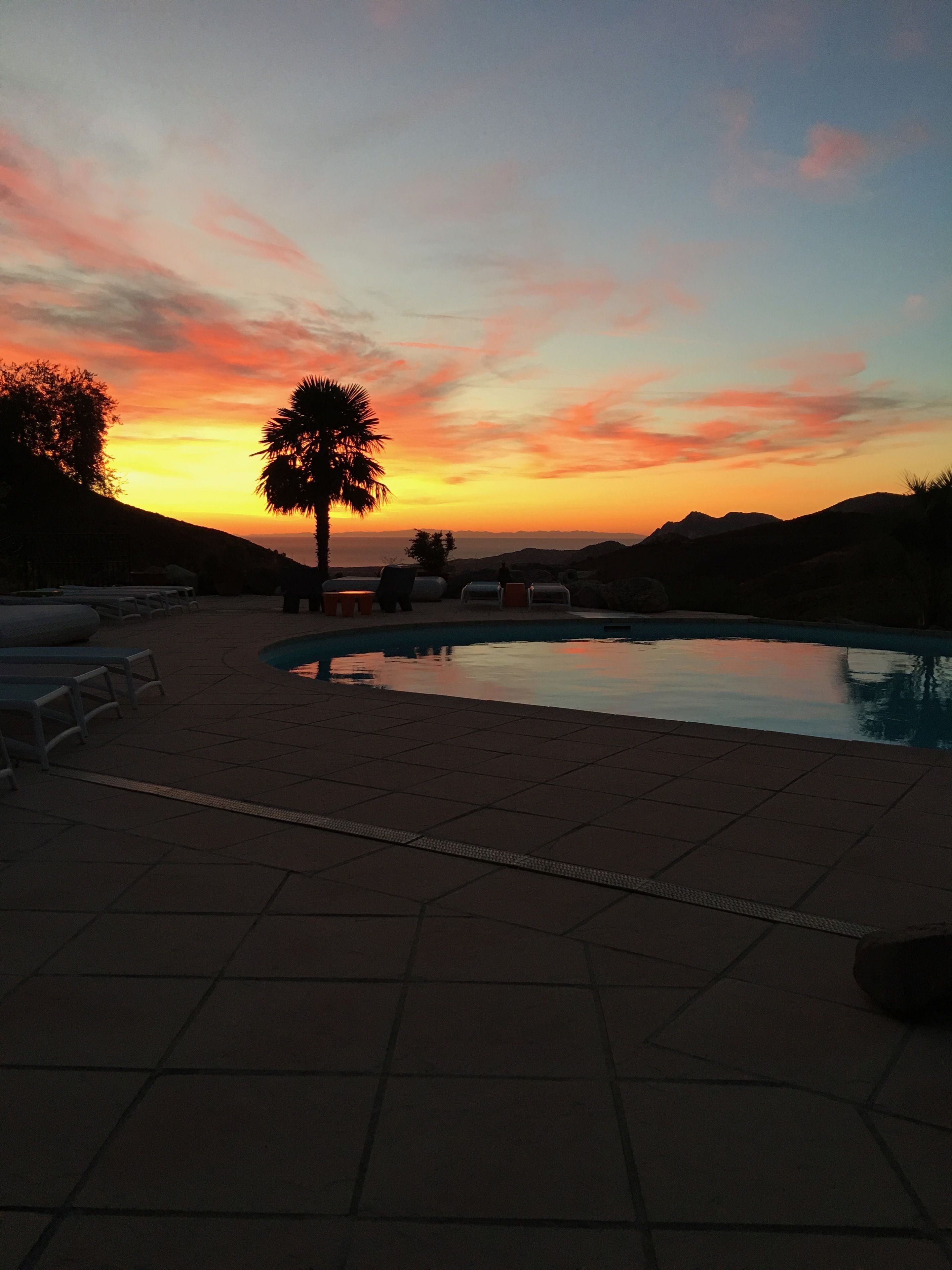 Uma área de piscina com espreguiçadeiras e uma palmeira, cercada por uma paisagem montanhosa ao pôr do sol.