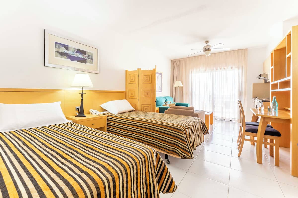 Quarto de hotel com duas camas, uma escrivaninha, uma cadeira, um guarda-roupa e um ventilador de teto.