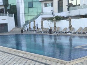 Piscina com espreguiçadeiras e guarda-sóis, uma escadaria e um edifício moderno com janelas de vidro.
