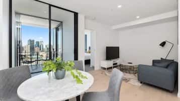 Uma sala de estar moderna com vista para a cidade, uma mesa redonda, cadeiras cinzas, um sofá cinza e uma televisão de tela plana.
