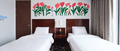 Quarto de hotel com duas camas, um chão com padrão geométrico, um mural floral na parede, uma mesinha de cabeceira e uma janela com cortinas.