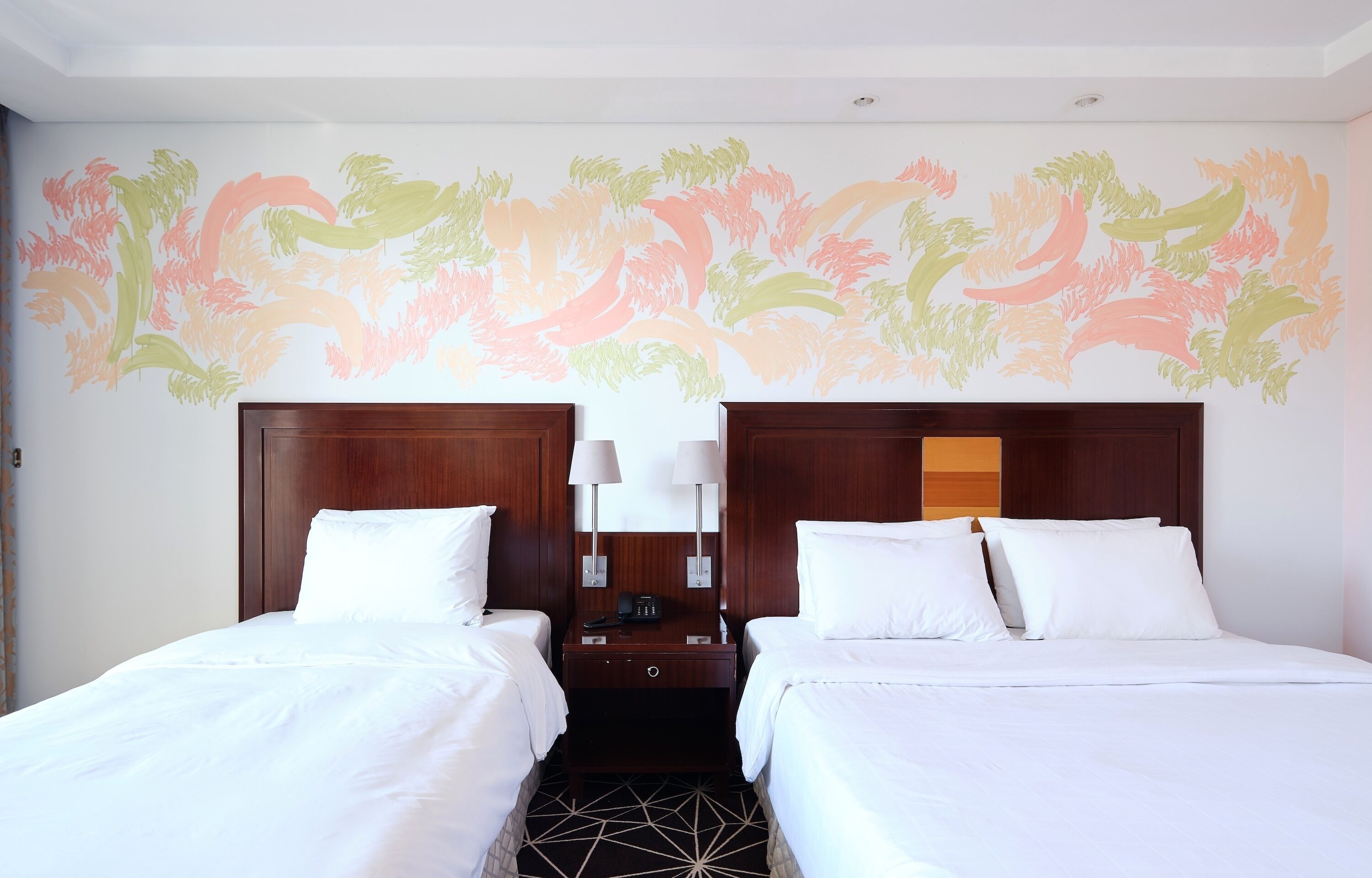 Quarto de hotel com duas camas, cabeceira de madeira, mesa de cabeceira com abajur e um mural na parede.