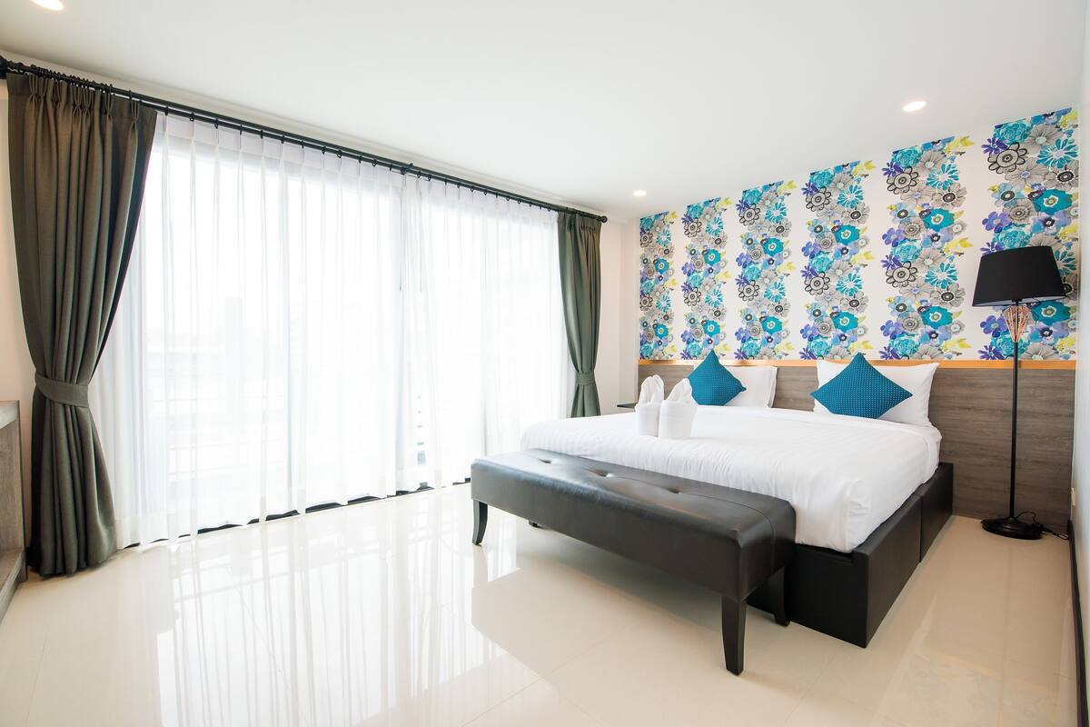 Quarto de hotel moderno com uma cama grande, um banco, uma luminária de parede e uma janela ampla com cortinas translúcidas.