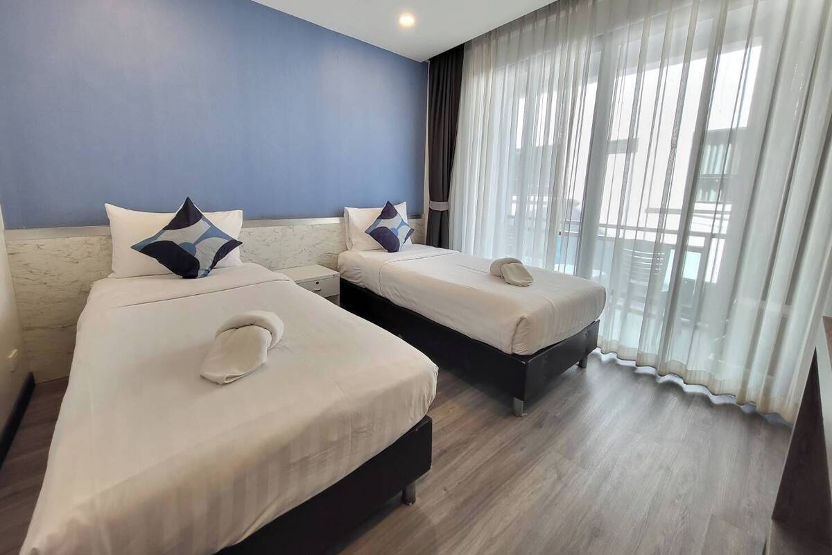 Quarto de hotel com duas camas, cabeceira de mármore, piso de madeira e uma janela grande com cortinas translúcidas.