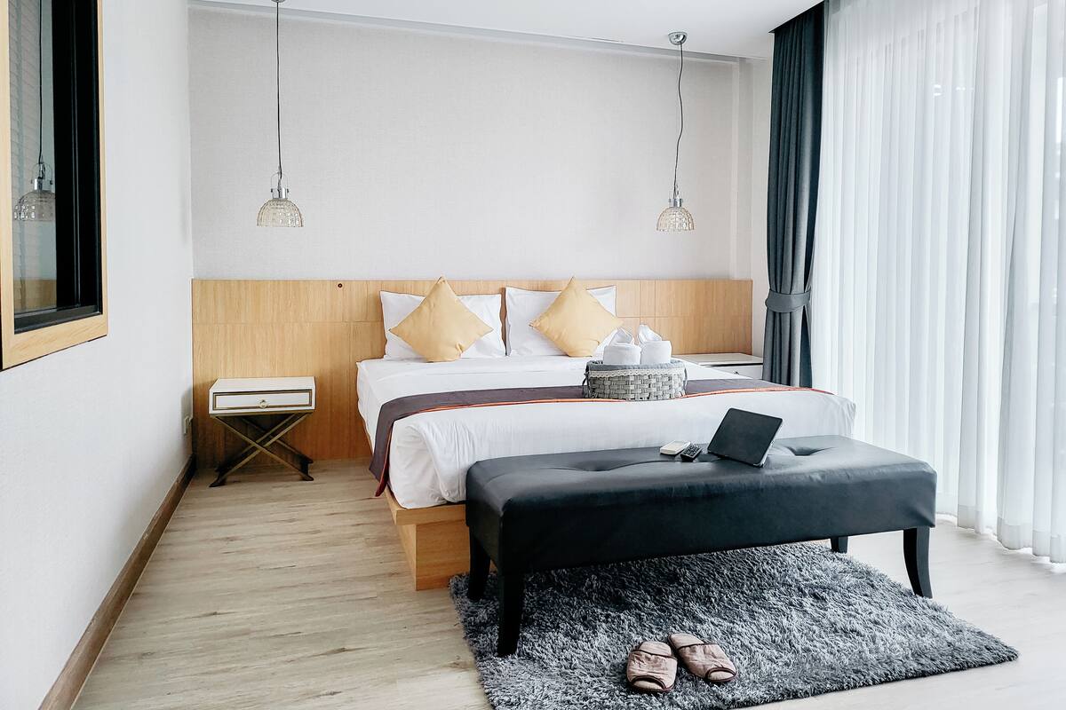 Quarto de hotel moderno com cabeceira de madeira, uma cama grande com roupa de cama branca, duas mesas de cabeceira, um abajur pequeno, um tapete cinza e um banquinho preto.