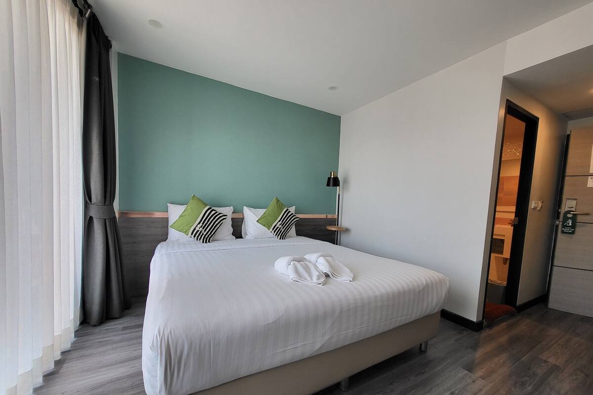 Quarto de hotel com uma cama grande, uma mesa de cabeceira, uma luminária e vista para o corredor.