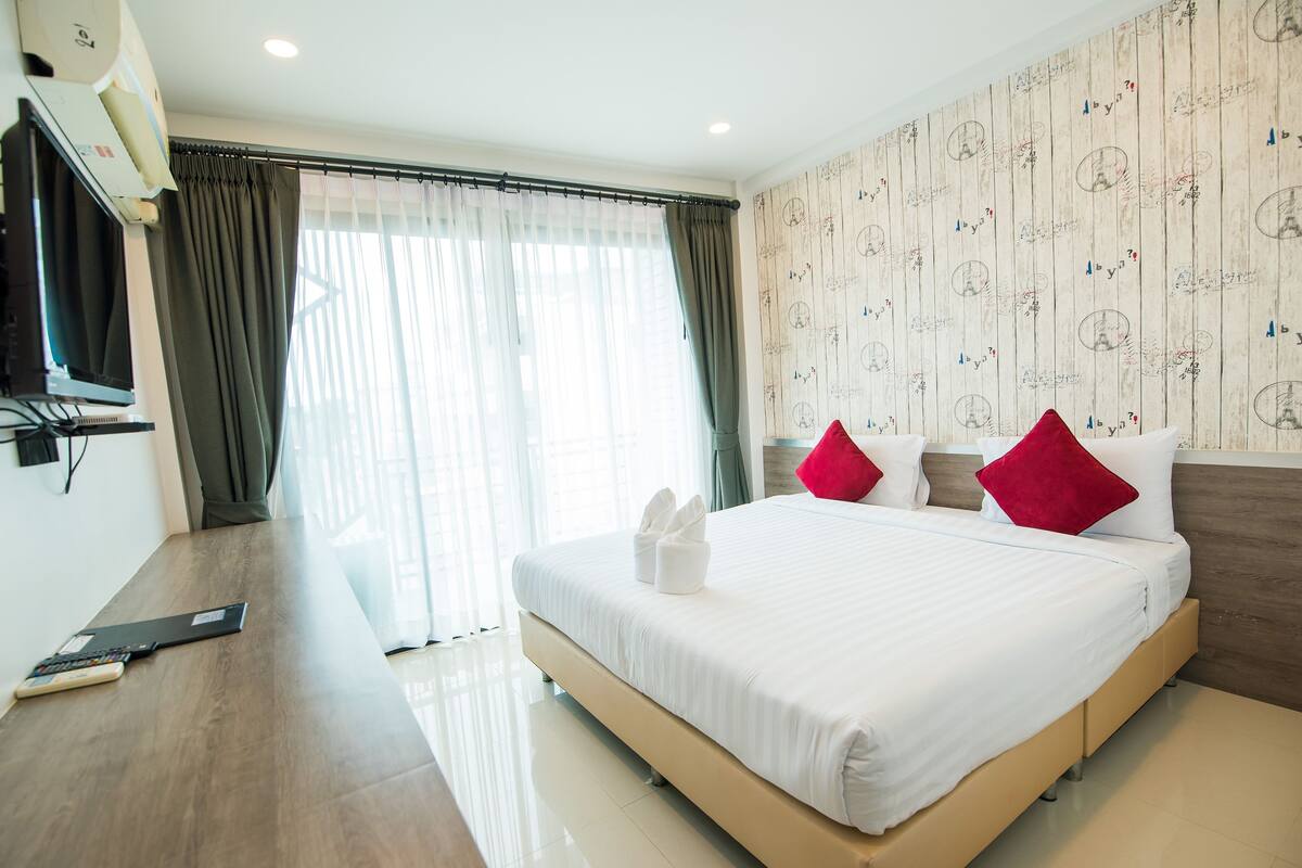 Quarto de hotel com uma cama grande, uma escrivaninha com um laptop, uma televisão e uma janela com cortinas.