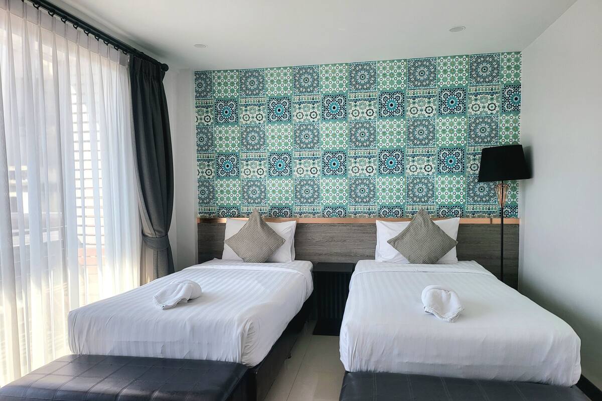 Quarto de hotel com duas camas, tapeçaria estampada, luminária de chão e uma janela grande com cortinas.