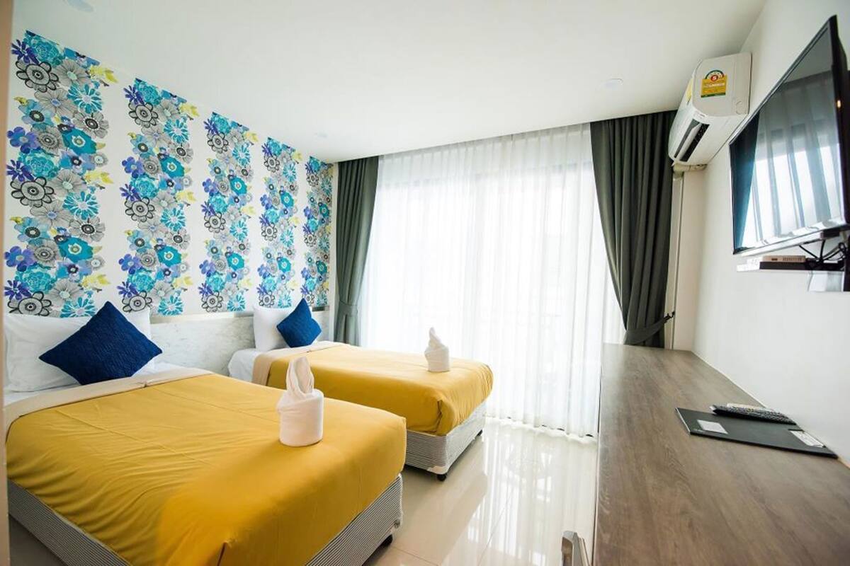 Quarto de hotel com duas camas, uma escrivaninha, ar condicionado e uma janela grande com cortinas.