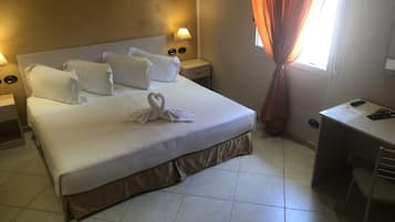 Cama bem arrumada com lençóis brancos e toalhas dispostas em formato de coração, uma escrivaninha com cadeira e uma janela com cortinas.