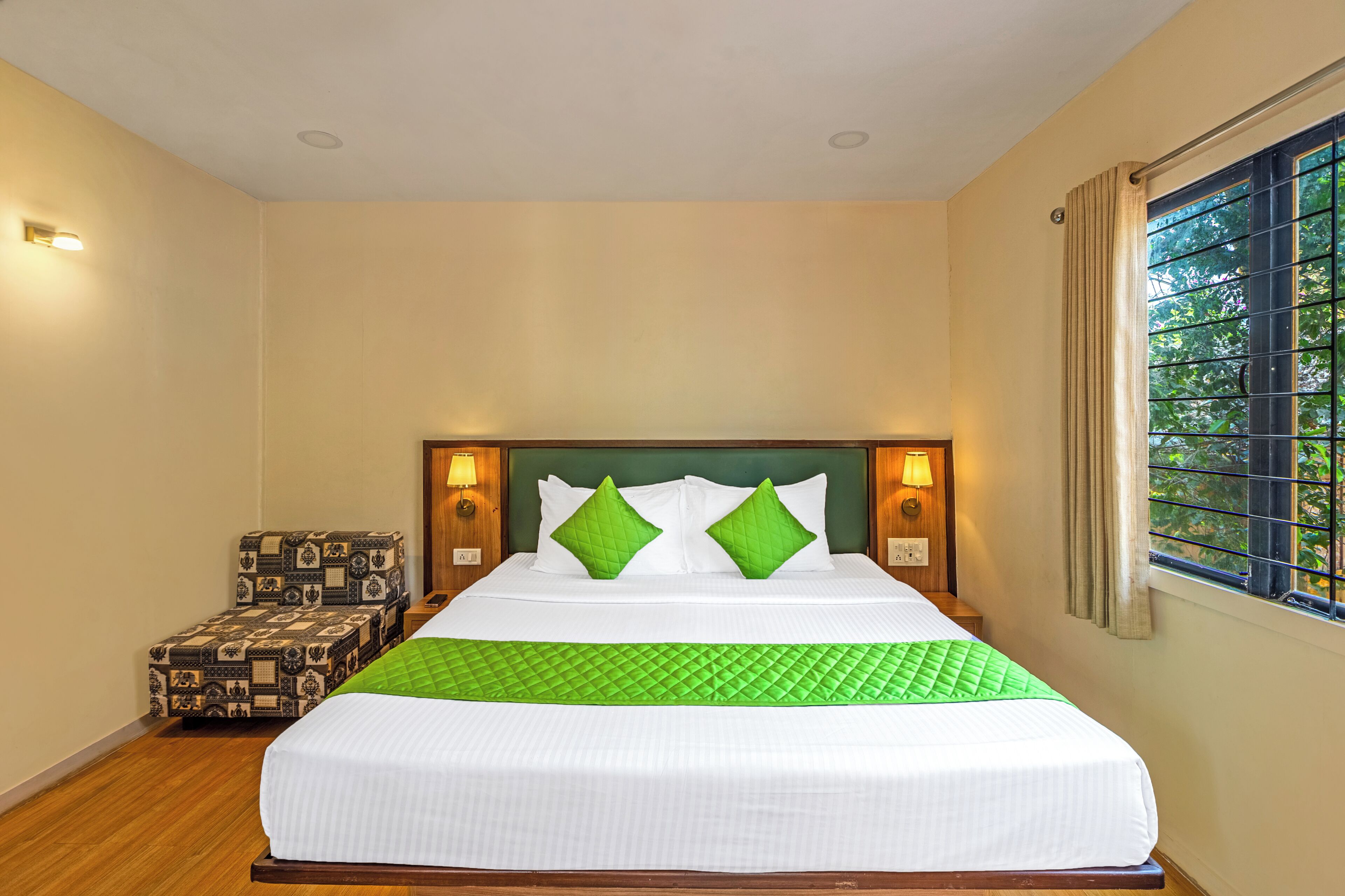 Quarto de hotel com uma cama grande, duas luminárias de cabeceira, uma janela com cortinas, uma cadeira pequena e piso de madeira.