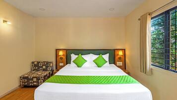 Quarto de hotel com uma cama grande, duas luminárias de cabeceira, uma janela com cortinas, uma cadeira pequena e piso de madeira.
