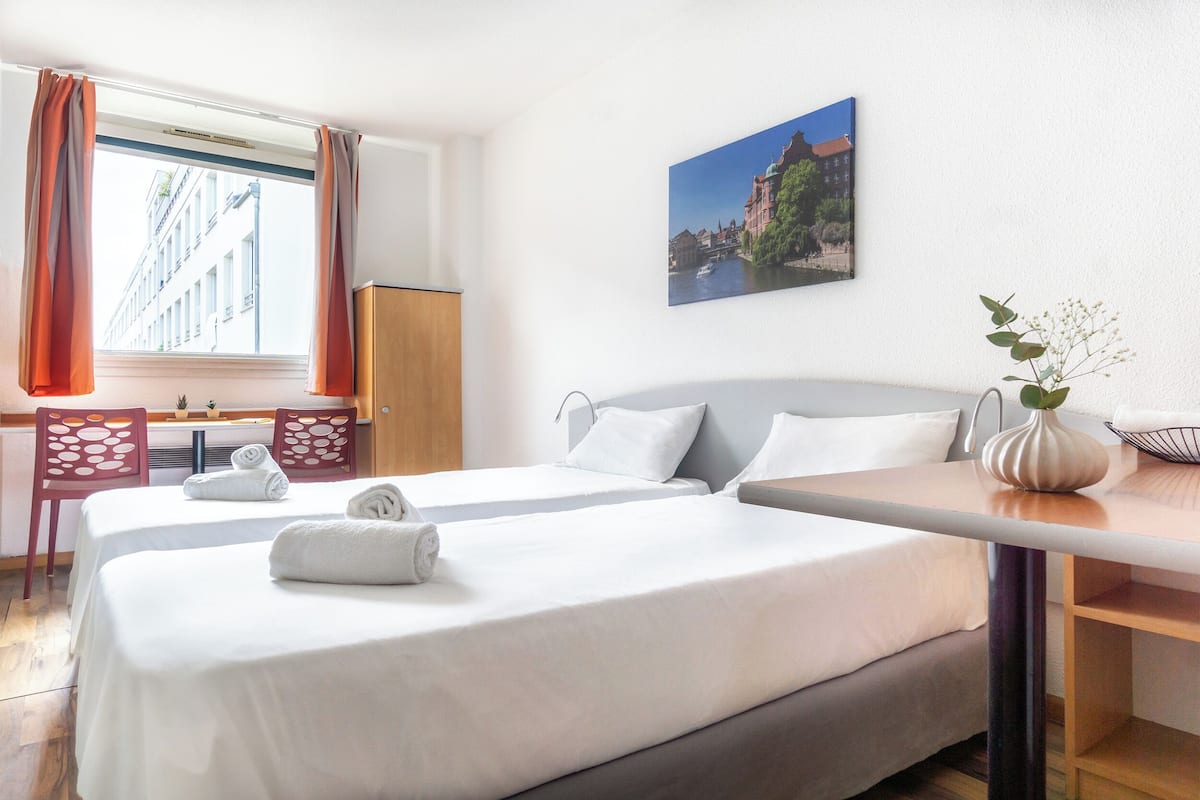 Quarto de hotel com duas camas, uma escrivaninha, cadeiras, um vaso com flores e um quadro na parede.