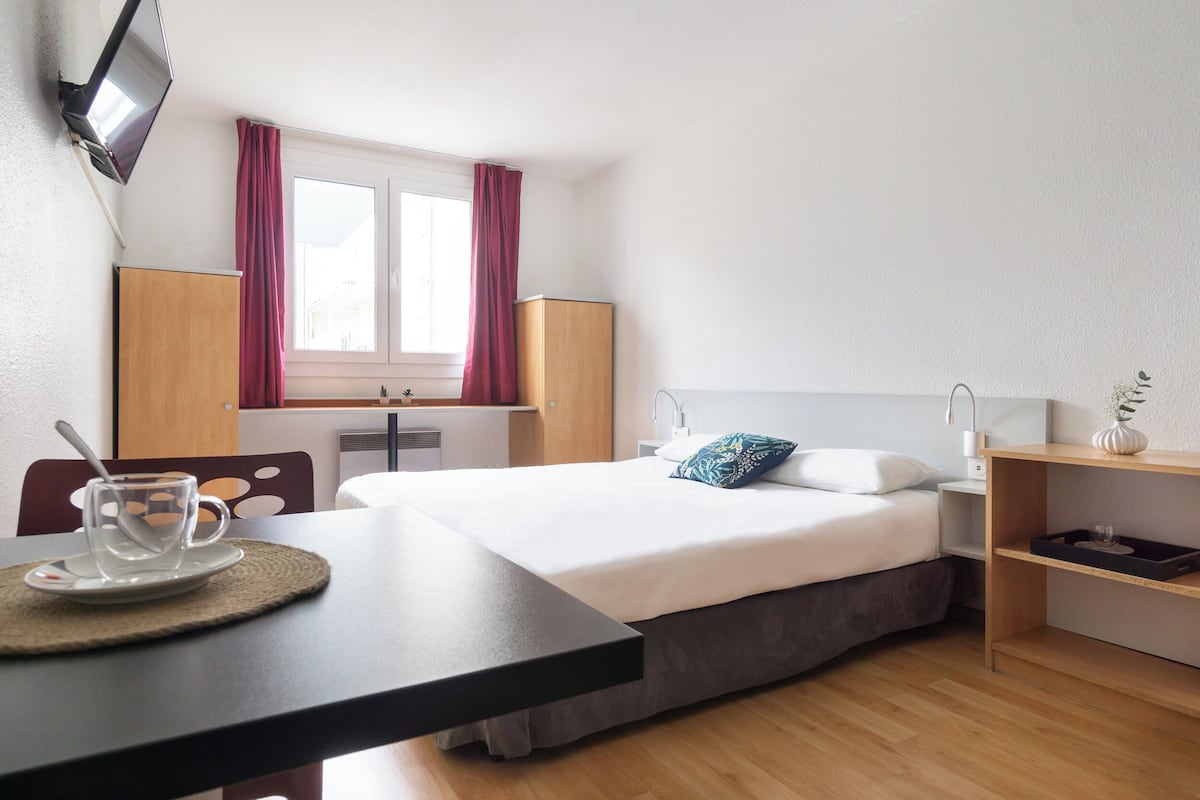 Quarto de hotel com cama, escrivaninha, cadeira, televisão e janela com cortinas.