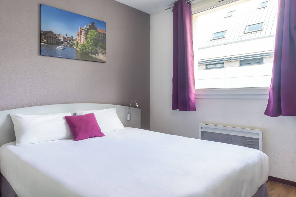 Quarto de hotel com cama, janela com cortinas roxas, quadro na parede com vista urbana e cabeceira branca.