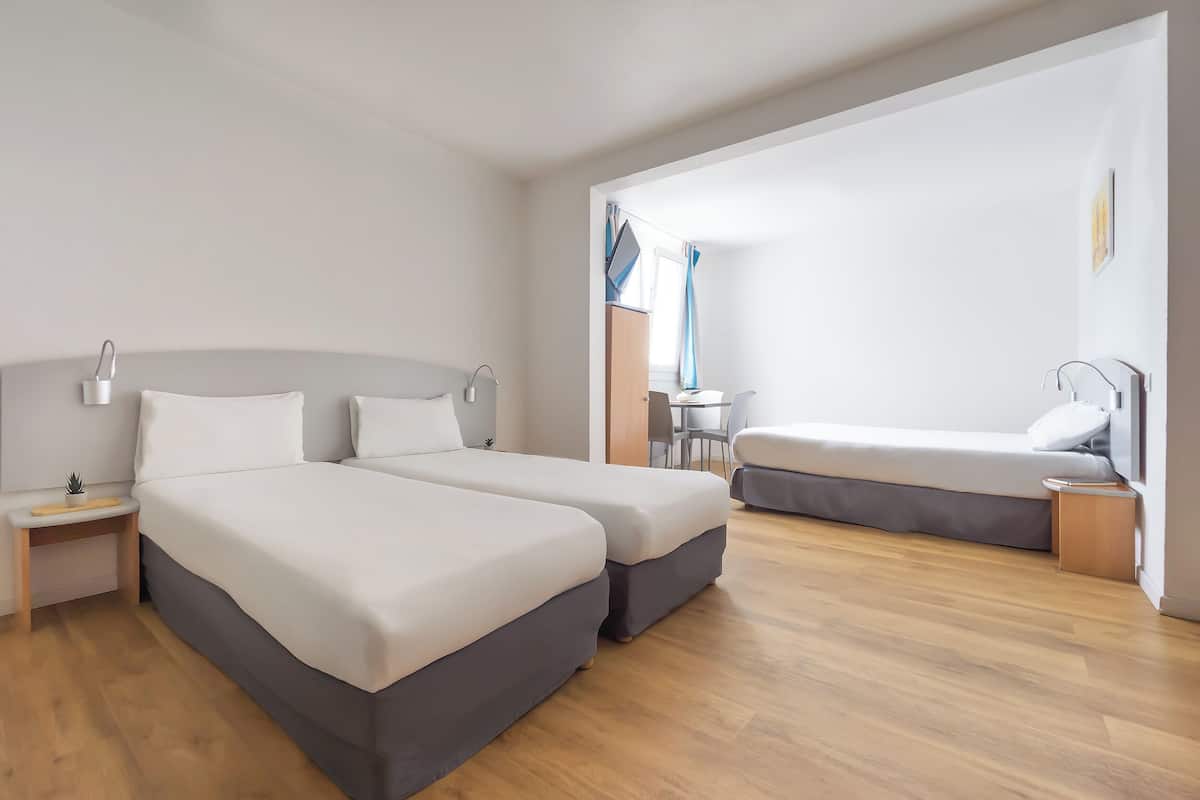 Quarto de hotel moderno com duas camas, piso de madeira e uma pequena escrivaninha com luminária.