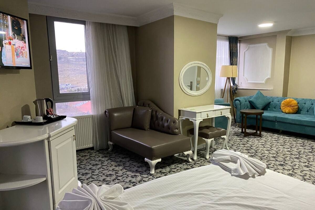 Quarto de hotel com cama, sofá, escrivaninha, espelho, janela com cortinas e vista para o exterior.
